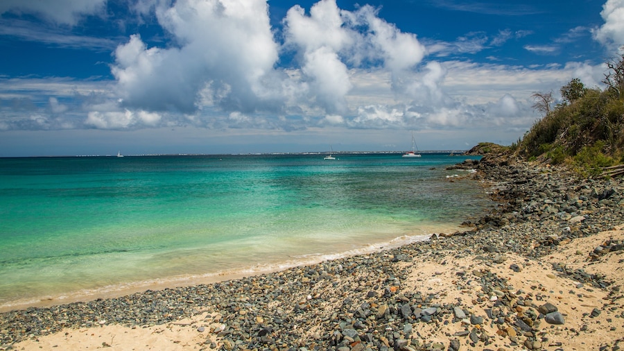 Uma praia de pedras com águas azul-turquesa e um veleiro ao longe.