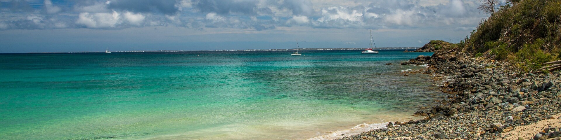 Uma praia de pedras com águas azul-turquesa e um veleiro ao longe.