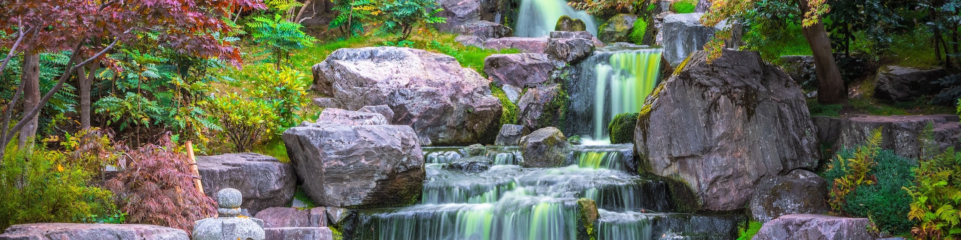Um jardim tranquilo com uma cascata, pedras grandes e diversas folhagens coloridas.