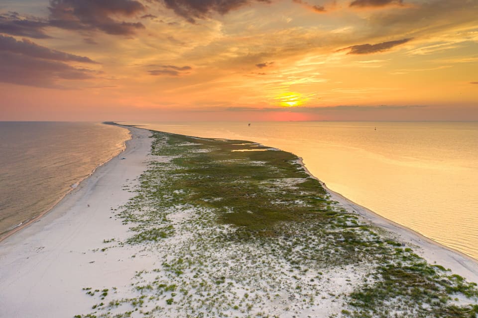 Uma praia com uma extensa faixa de areia e vegetação rala, que se estende até o oceano sob um céu ao pôr do sol.