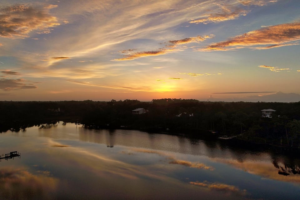 Um pôr do sol sobre um lago calmo com reflexos, rodeado por árvores e edifícios ao longe.