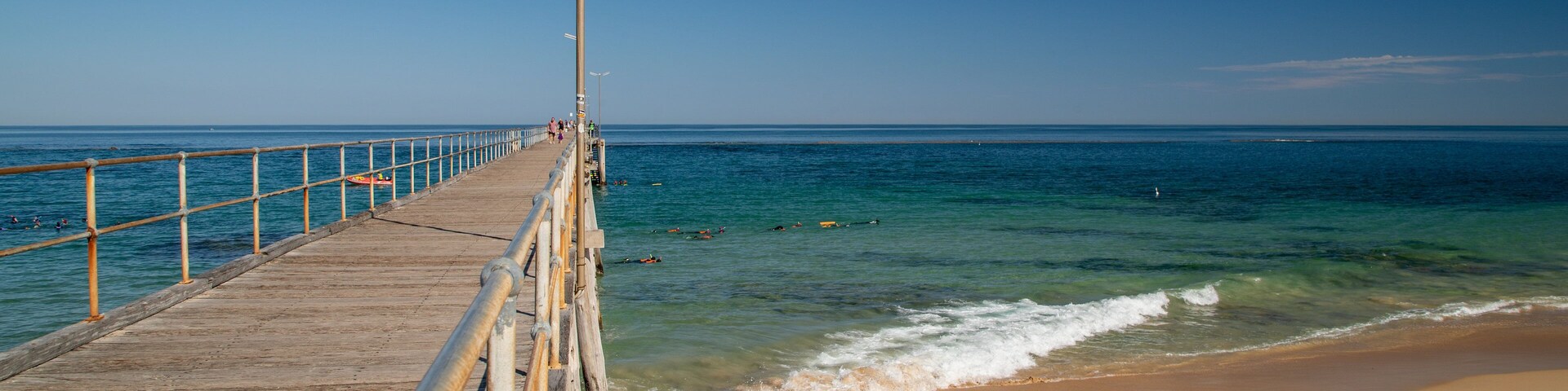Um cais de madeira que se estende para o oceano, com pessoas caminhando e um casal na praia.
