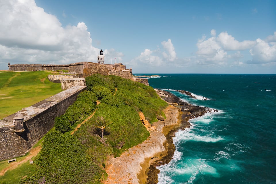 Uma fortificação costeira com farol, vegetação exuberante e o oceano.