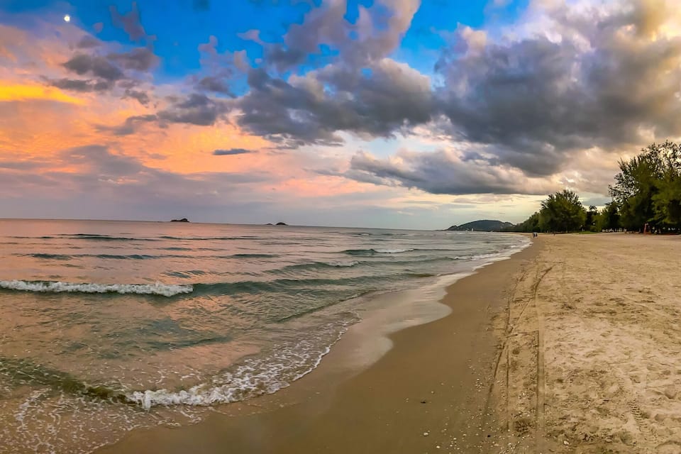 Uma praia com ondas, céu claro e montanhas ao longe.