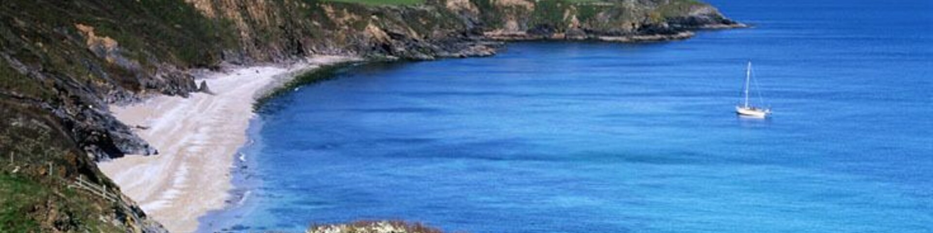 Paisagem costeira com praia de areia, mar azul límpido e falésia gramada.