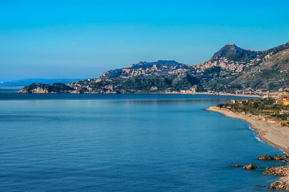 Uma vila costeira com montanhas ao fundo, mar calmo e costa rochosa.