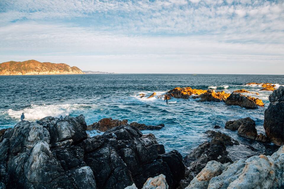 Litoral rochoso com ondas quebrando nas pedras e uma cadeia de montanhas distante sob um céu parcialmente nublado.