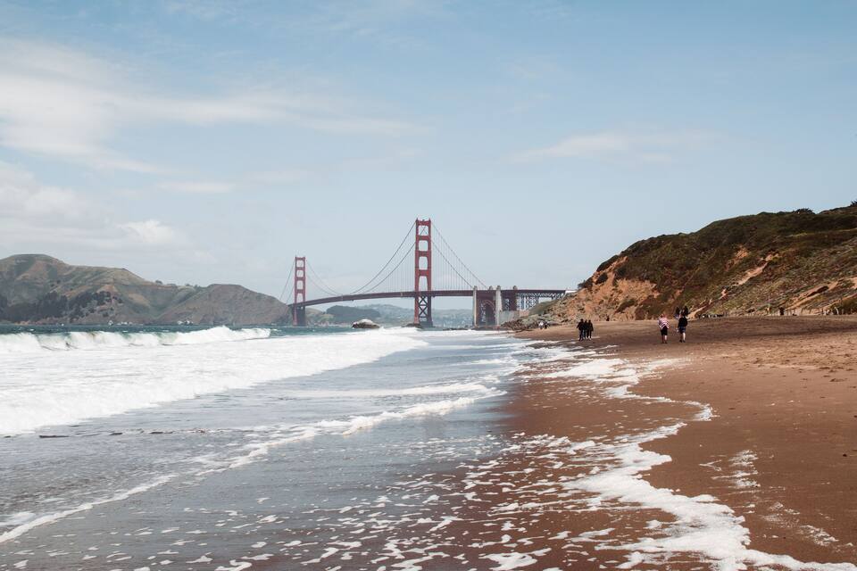 Uma praia com ondas, uma ponte vermelha e pessoas caminhando.