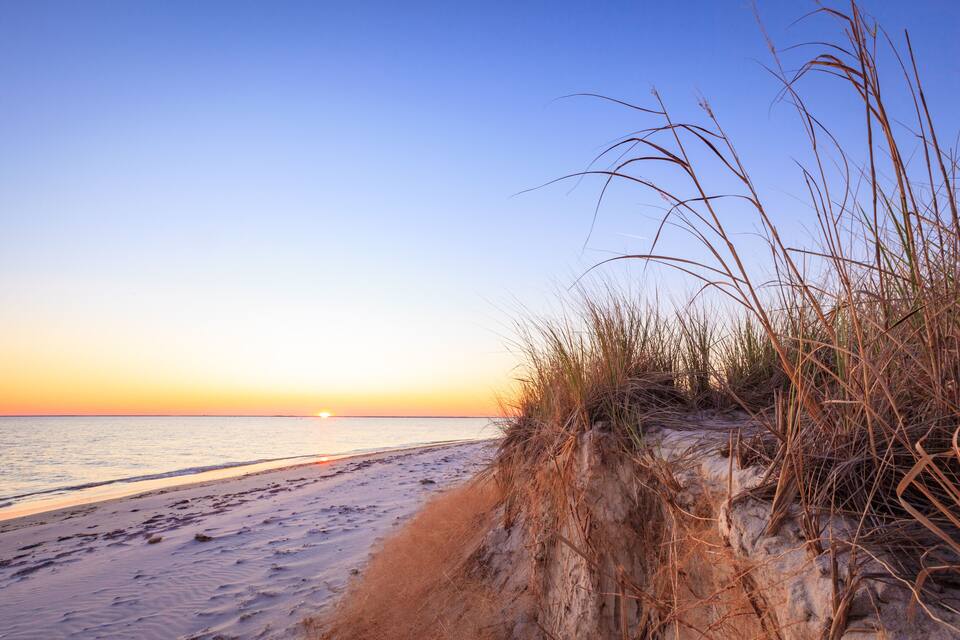 Uma praia ao pôr do sol com dunas de areia e gramíneas altas.