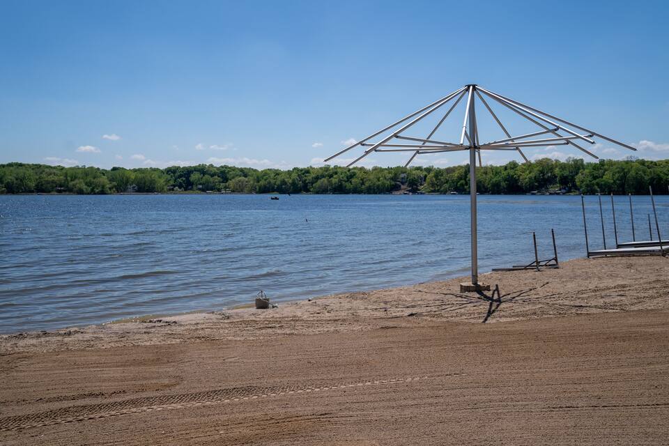 Uma praia com uma estrutura de guarda-sol de metal, águas calmas e uma orla arborizada.