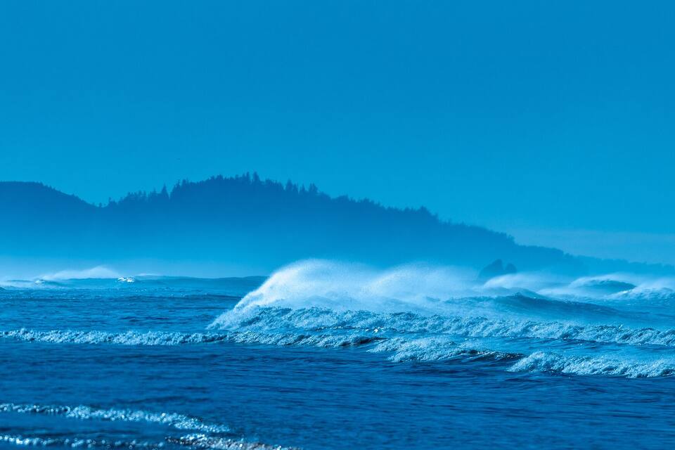 Um oceano extenso com ondas quebrando na costa e uma cadeia de montanhas ao longe.