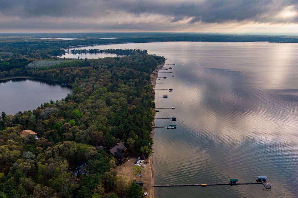 Vista aérea de uma área à beira de um lago, com um píer que se estende sobre a água, rodeada por uma floresta densa e um pequeno povoado com casas e barcos.