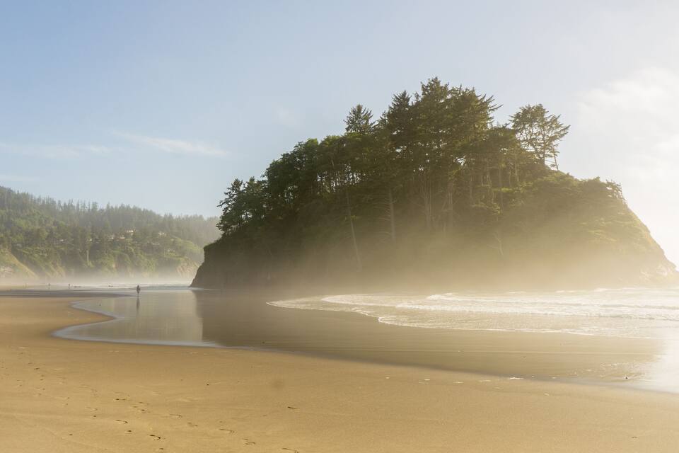 Uma praia arenosa com uma grande formação rochosa coberta de árvores, rodeada por colinas arborizadas.