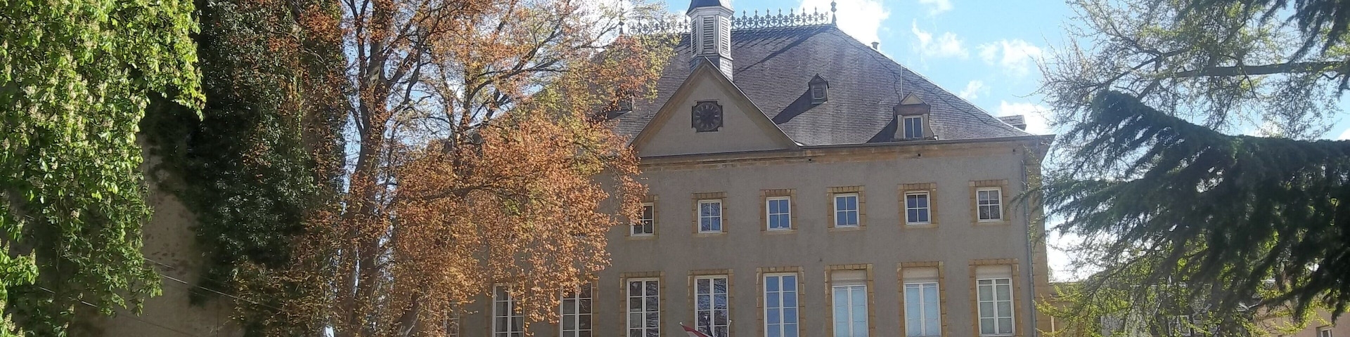 Um edifício grande e histórico com uma torre do relógio, rodeado por jardins e árvores bem cuidados.