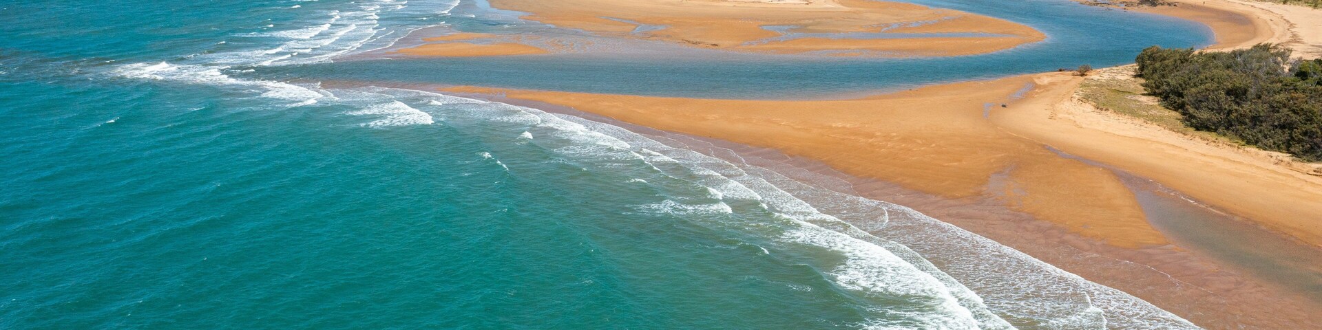 Paisagem costeira com uma praia arenosa, águas turquesa e um rio serpenteando pelo terreno.