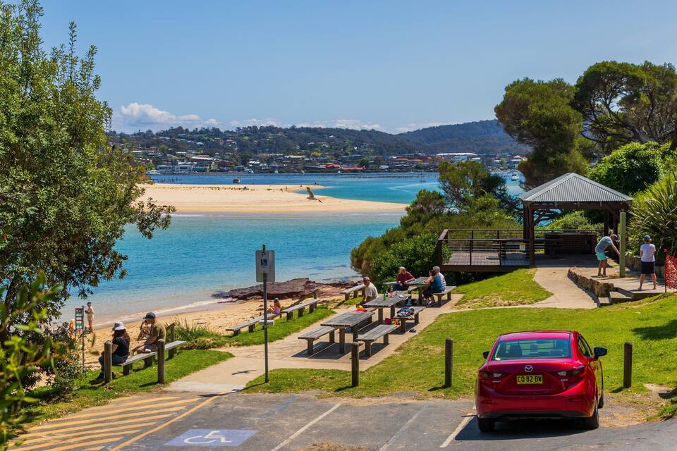 Um parque costeiro com áreas de piquenique, um carro vermelho e uma praia ao fundo.