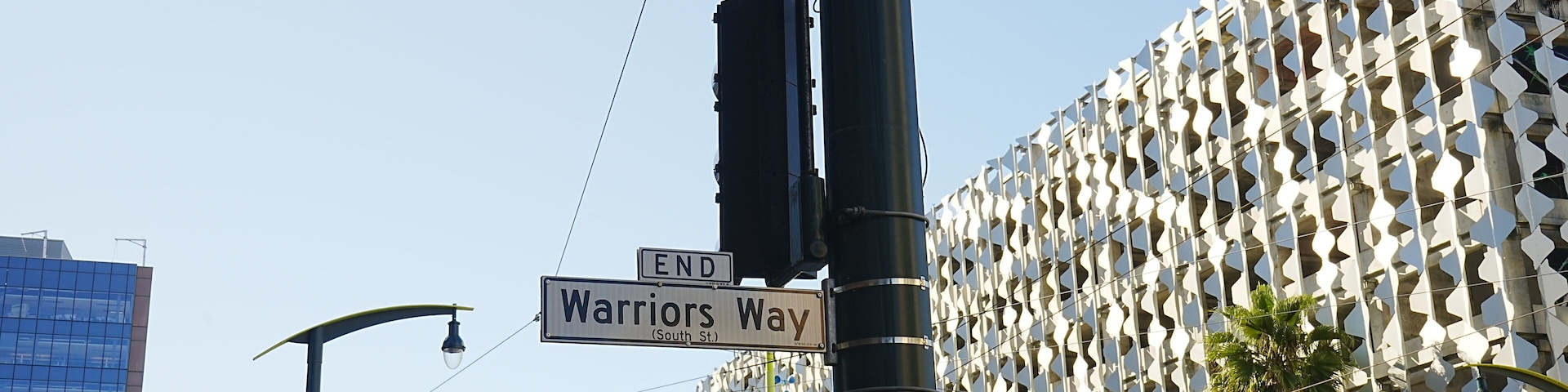 Placa de rua indicando 'Warriors Way (South St)' com um edifício moderno e fachada geométrica distinta ao fundo.