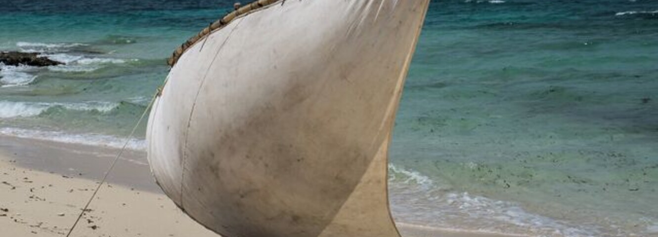 Um veleiro tradicional numa praia de areia, águas turquesa cristalinas e uma linha de costa distante.