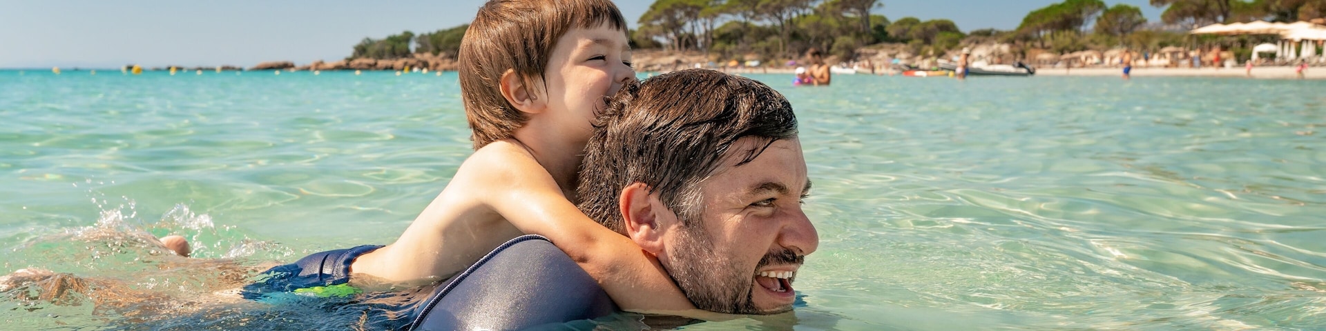 Um homem carregando uma criança nas costas em água azul cristalina, com uma praia e árvores ao fundo.