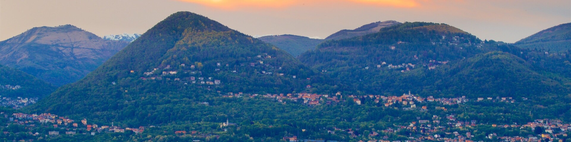 Paisagem montanhosa com uma cidade na base, um corpo d'água em primeiro plano e um pôr do sol ao fundo.