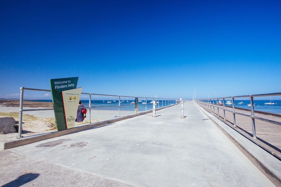 Uma passarela de concreto com uma placa indicando 'Welcome to Flinders Jetty', levando a uma praia com barcos na água.