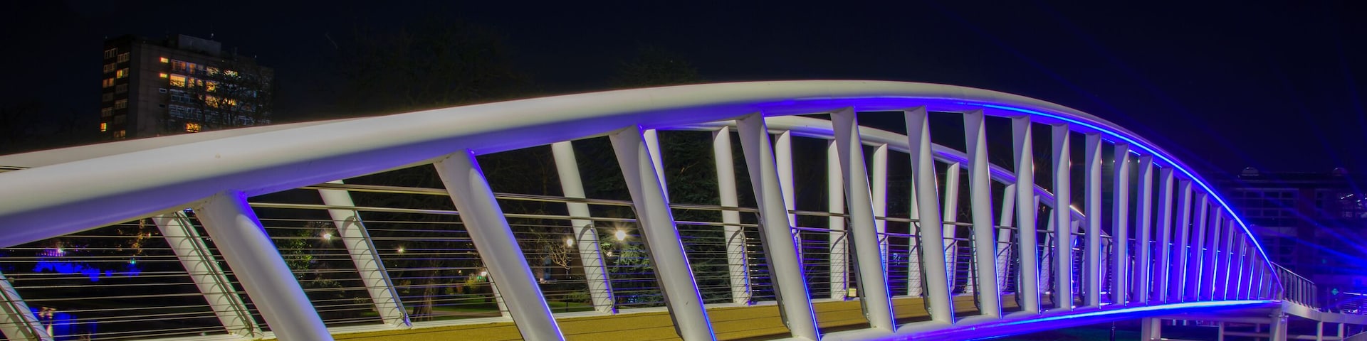 Uma ponte moderna com guarda-corpos brancos e iluminação azul, atravessando um corpo de água com um horizonte urbano ao fundo.