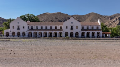 Um edifício histórico com janelas em arco e telhado ladrilhado, cercado por um pátio de cascalho e montanhas ao fundo.