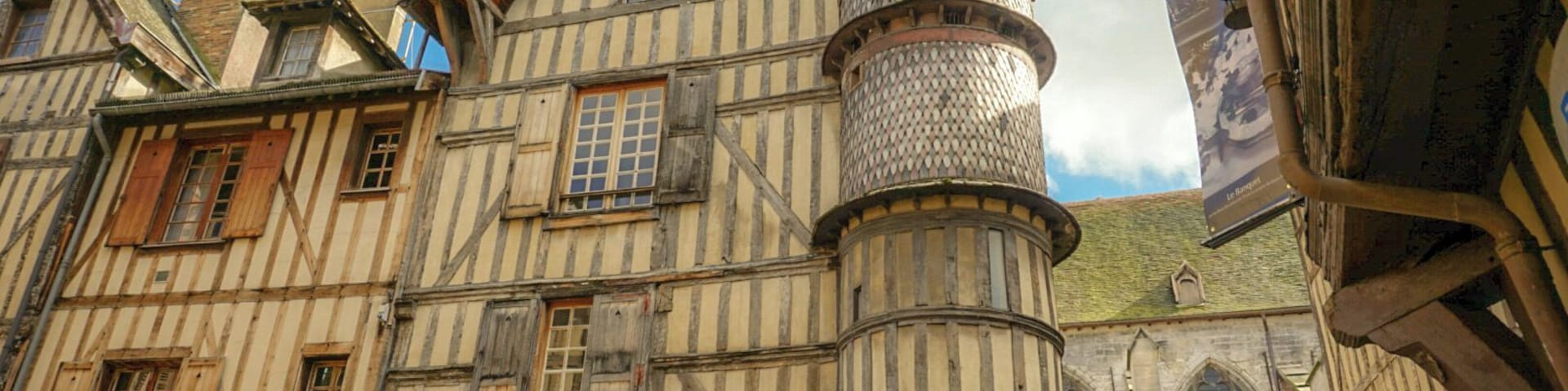 Um edifício histórico com estrutura de madeira, uma torre cilíndrica e uma igreja ao fundo.