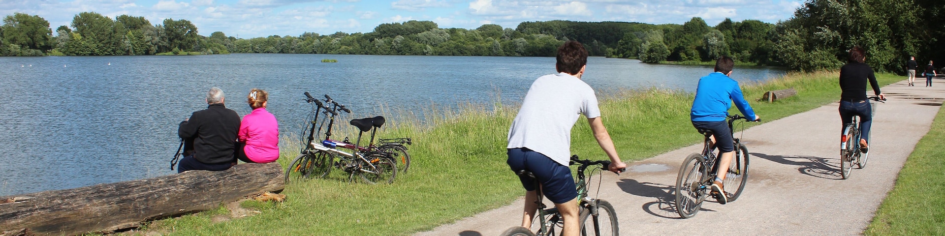 Um caminho à beira do lago com ciclistas e um casal sentado num tronco.