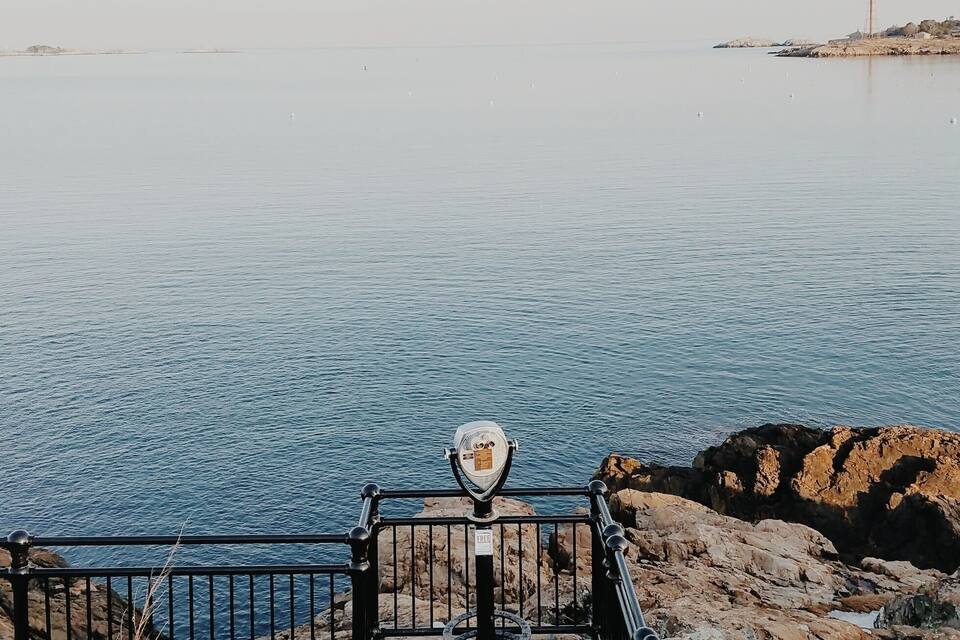 Cena costeira com um telescópio numa plataforma rochosa com vista para águas calmas.