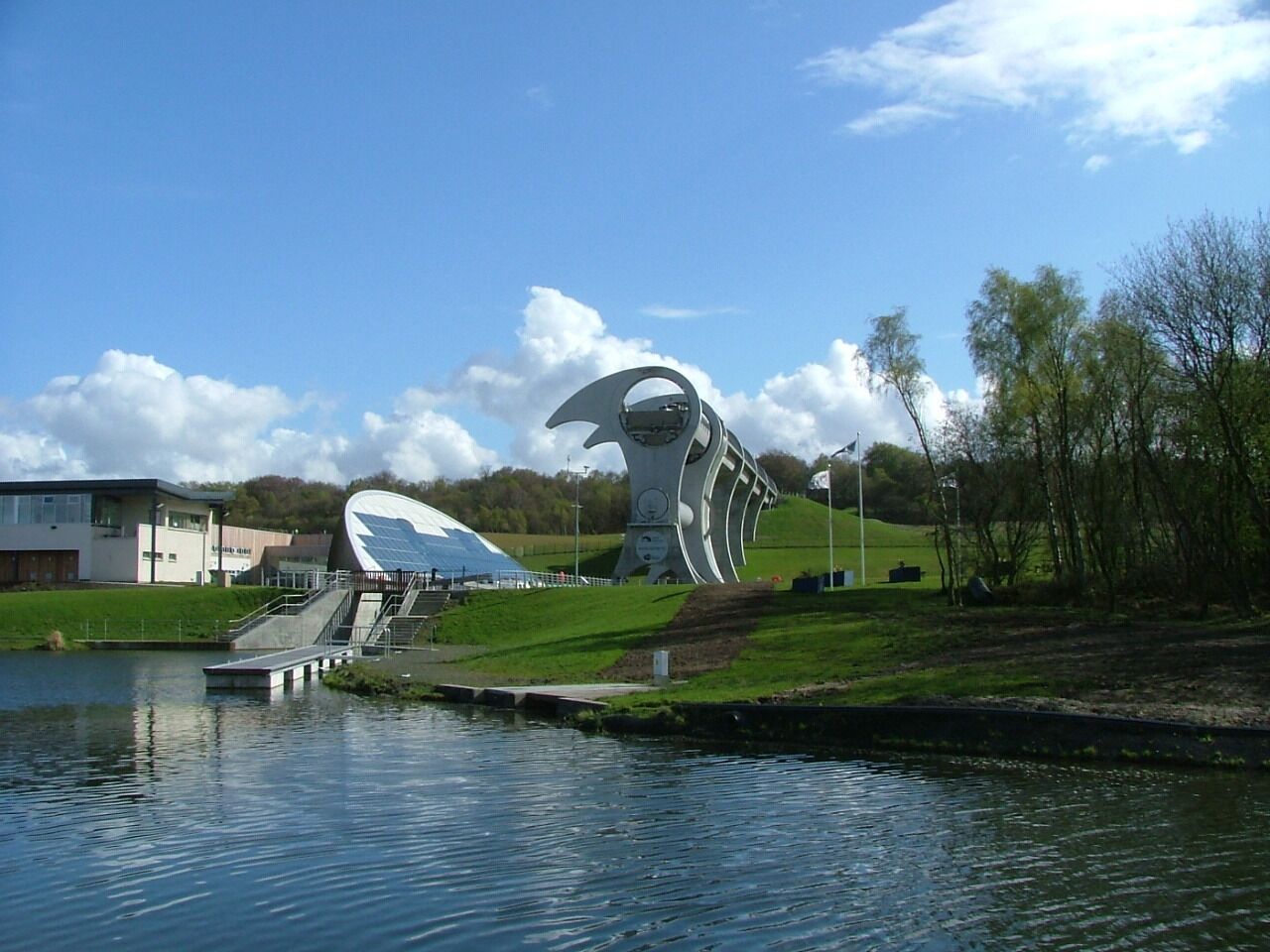 Uma escultura moderna perto da água, uma ponte e um edifício ao fundo.