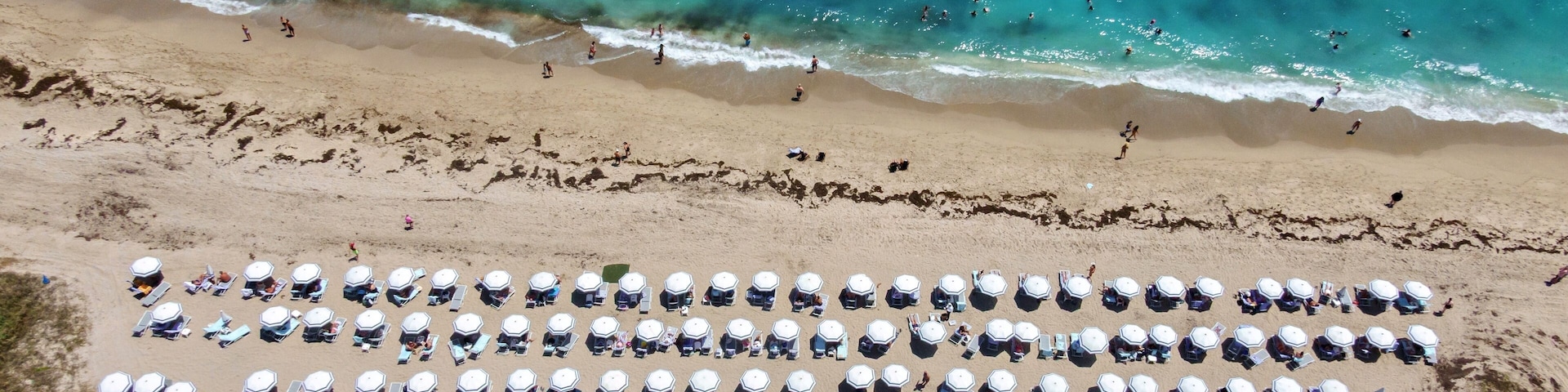 Vista aérea de uma praia com muitos guarda-sóis brancos e espreguiçadeiras dispostas em um padrão circular, com o oceano e ondas ao fundo.