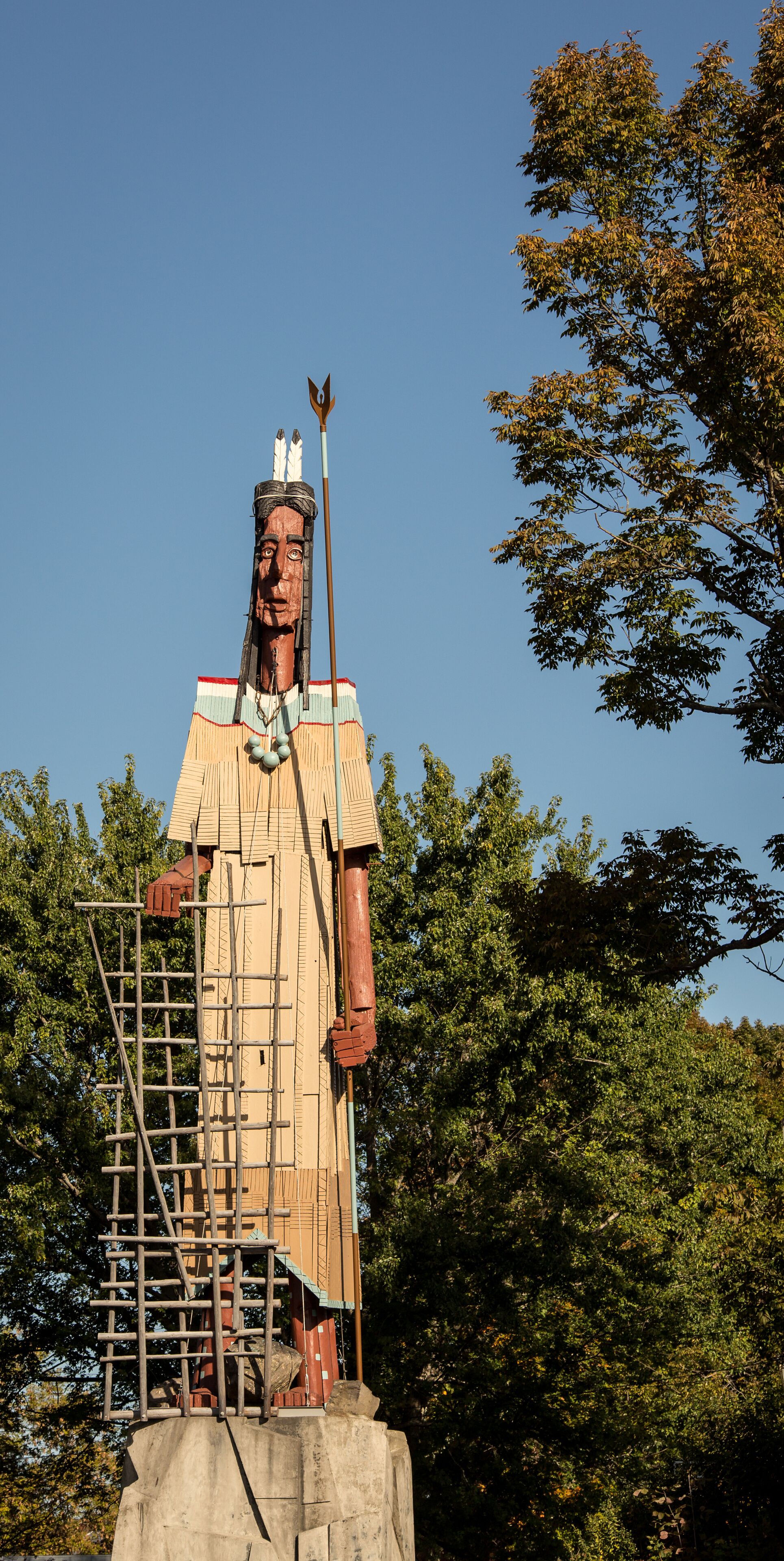 Uma grande estátua de uma figura nativo americano, com uma lança e escudo, rodeada por árvores.