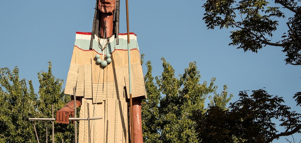 Uma grande estátua de uma figura nativo americano, com uma lança e escudo, rodeada por árvores.