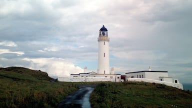 Um farol branco, com a parte superior azul, cercado por um muro branco e uma colina gramada.