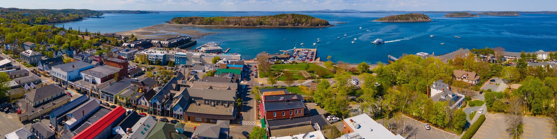Uma vila costeira com edifícios, uma marina e ilhas ao fundo.