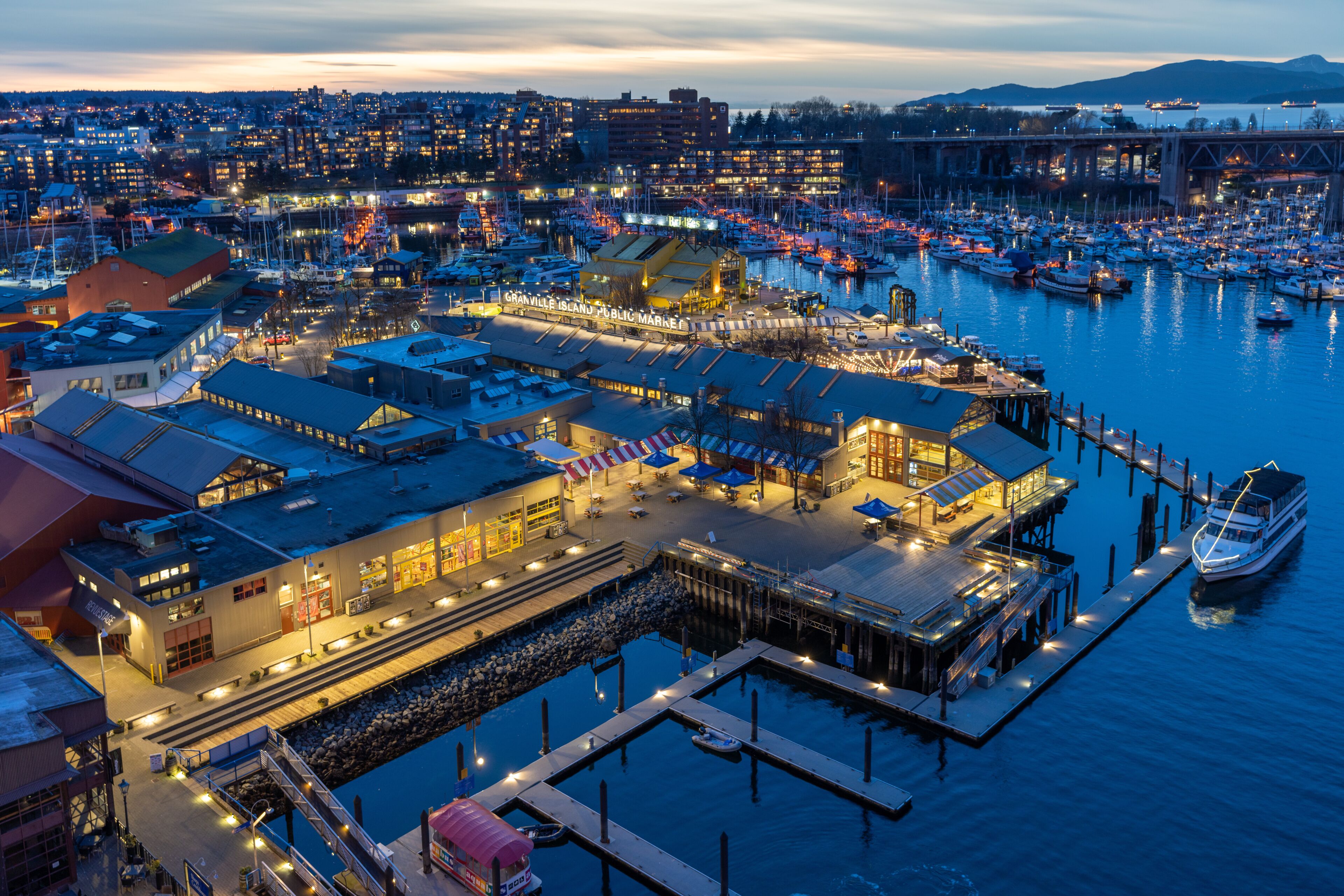 Uma marina com barcos, um cais e edifícios iluminados ao anoitecer.