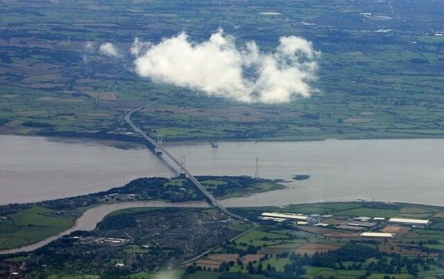 Uma ponte sobre um rio, cercada por campos verdes e uma pequena ilha com edifícios.