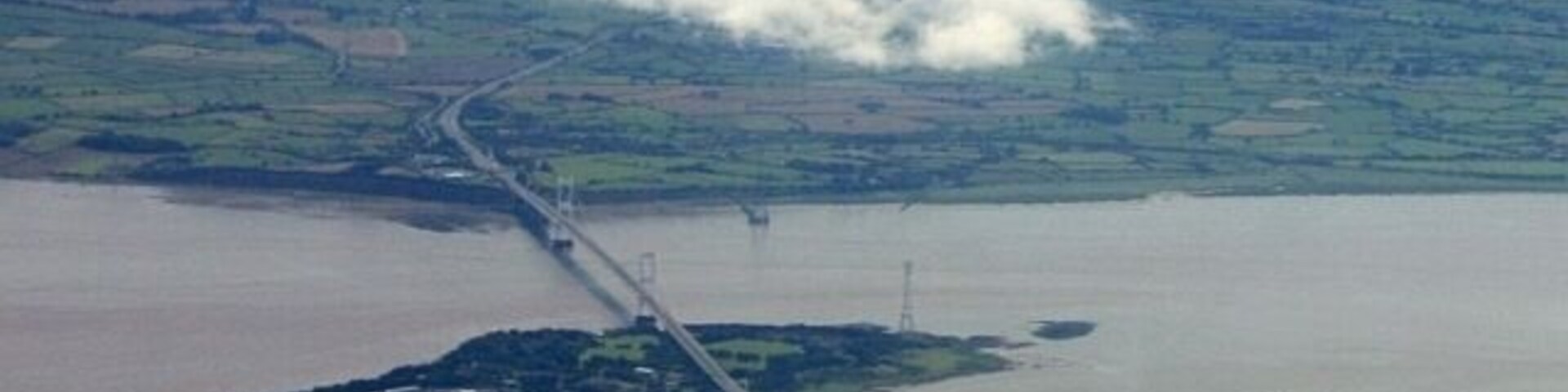 Uma ponte sobre um rio, cercada por campos verdes e uma pequena ilha com edifícios.