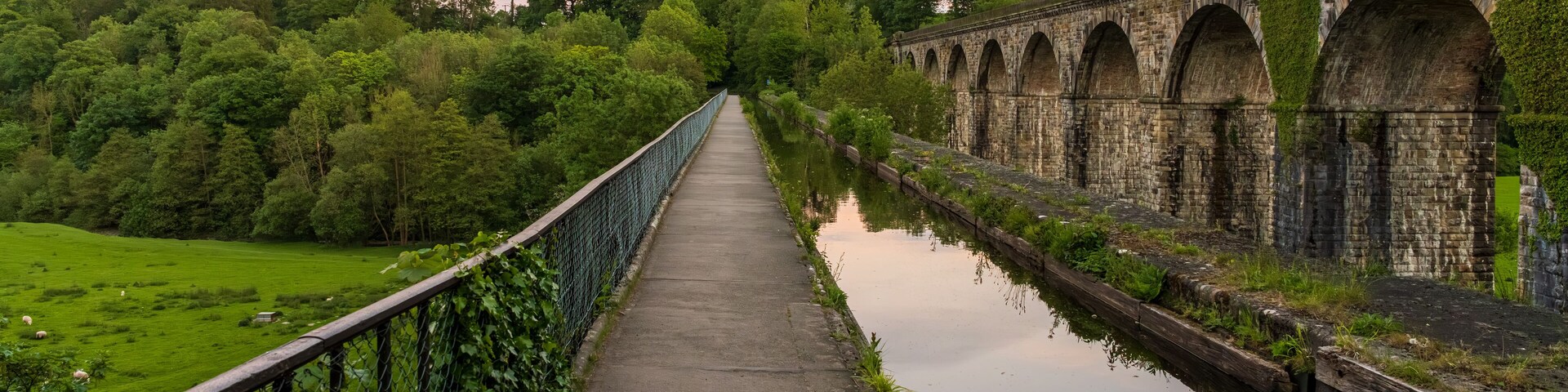 Uma ponte sobre um canal, com uma passarela de um lado e um campo gramado do outro.