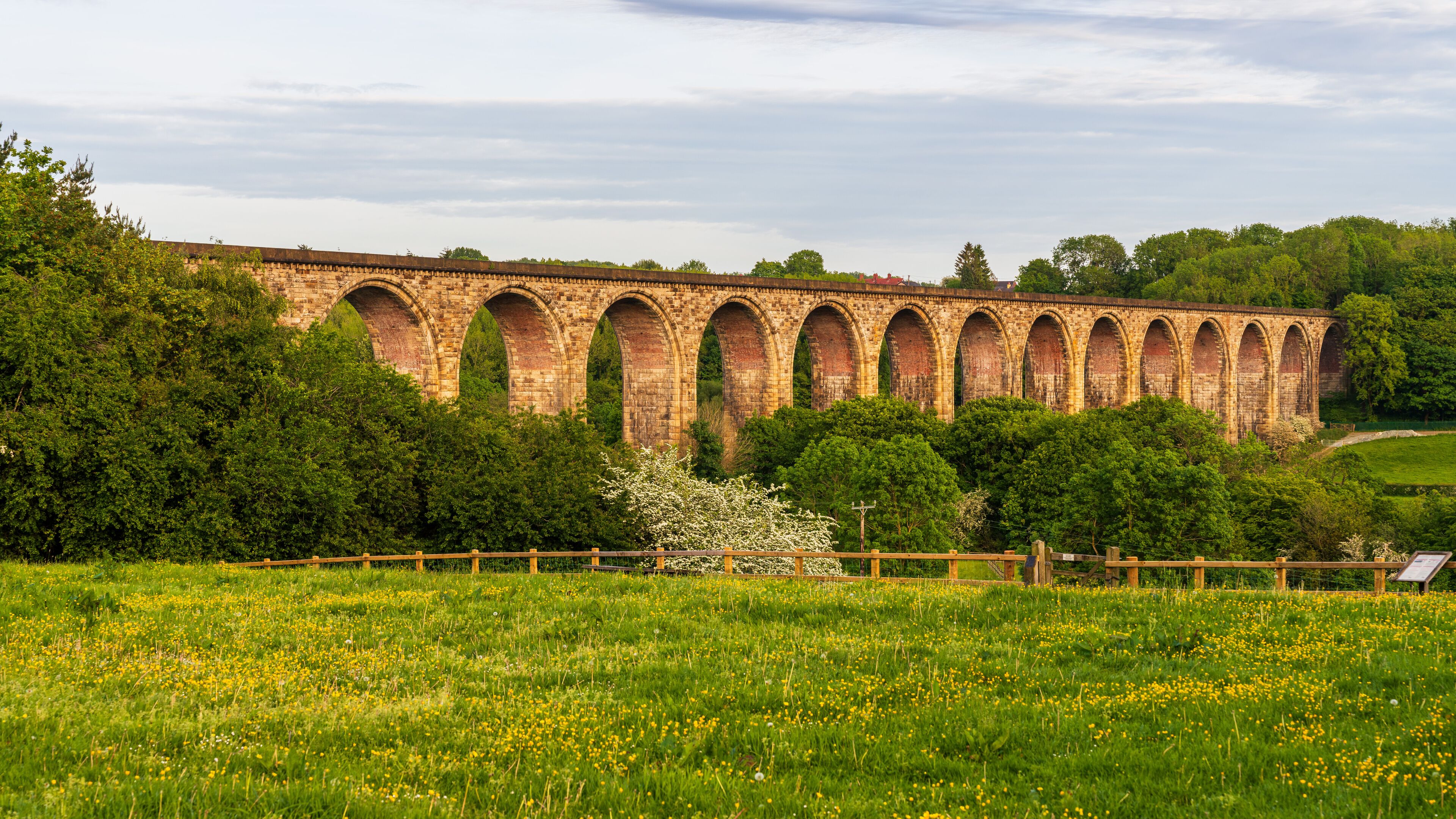 Um viaduto de pedra com múltiplas abóbadas, rodeado por vegetação exuberante e um campo de flores amarelas.