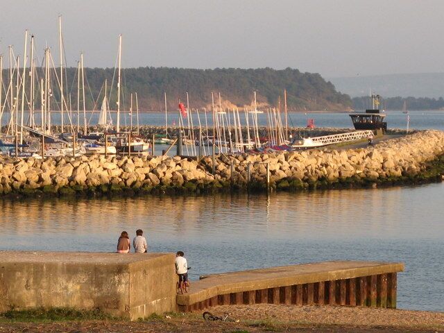 Uma marina com barcos, um paredão rochoso e uma colina arborizada ao fundo.