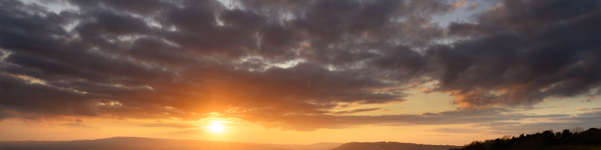 Um pôr do sol sobre uma paisagem acidentada com nuvens dispersas e uma aldeia lá embaixo.