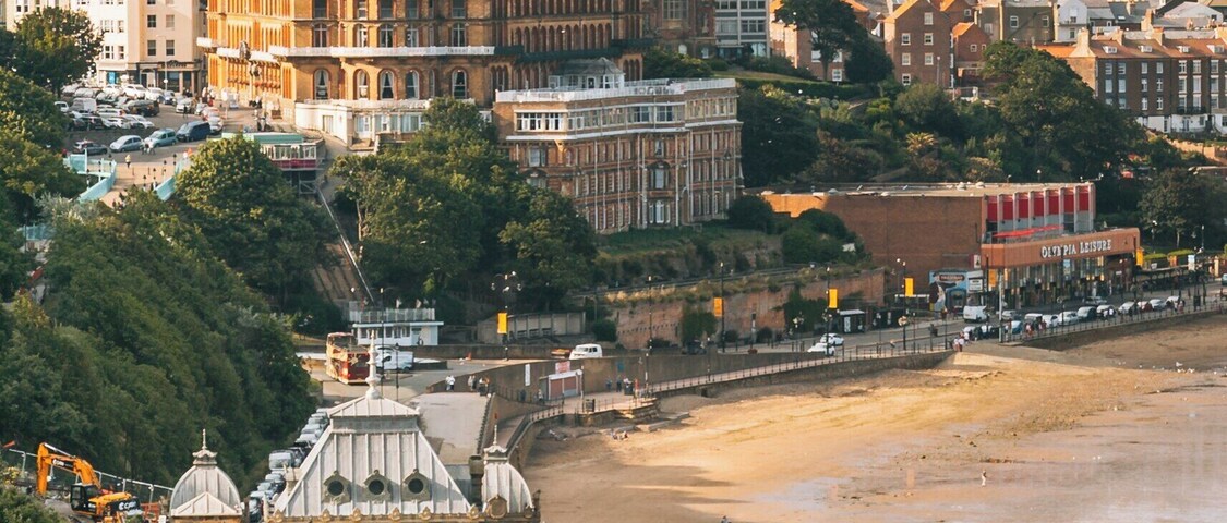 Paisagem costeira urbana com um edifício histórico imponente com cúpulas, uma praia de areia e um calçadão com carros estacionados.