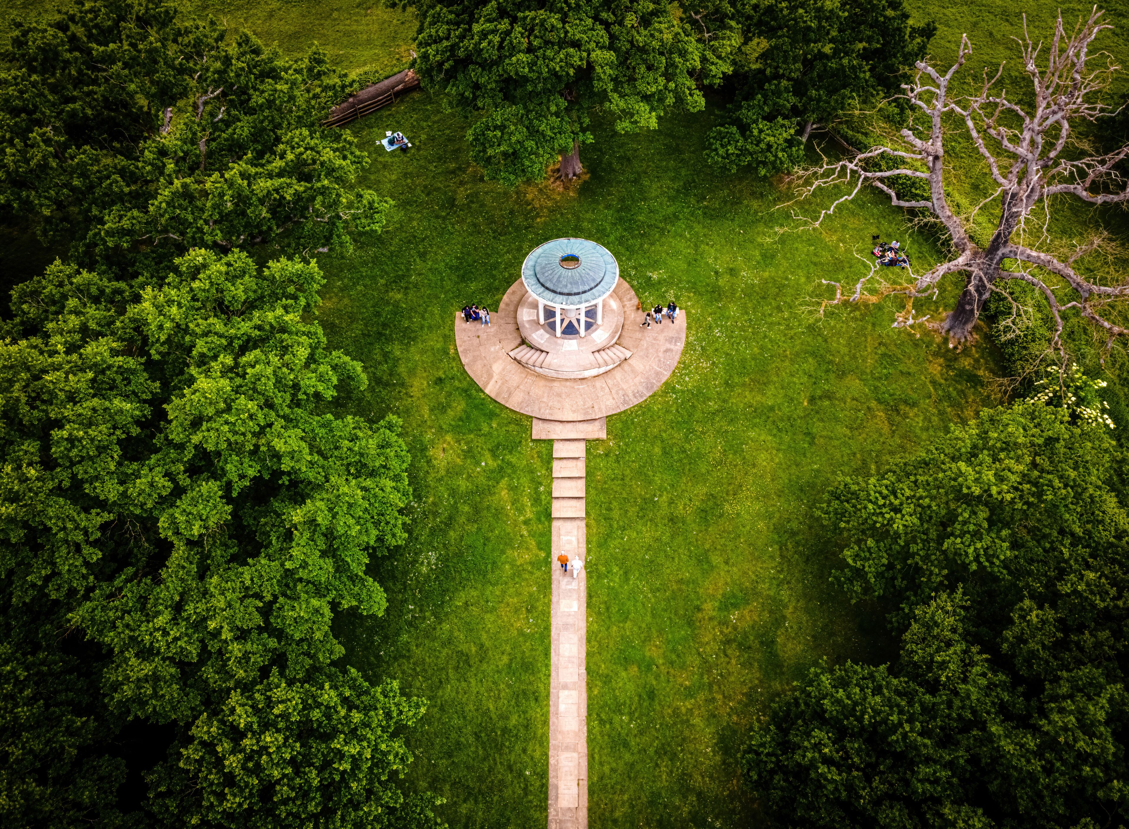 Vista aérea de um parque com um coreto central cercado por vegetação exuberante e uma árvore imponente.