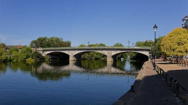 Uma ponte de pedra sobre um rio calmo, com árvores e um espaço com bancos à direita.