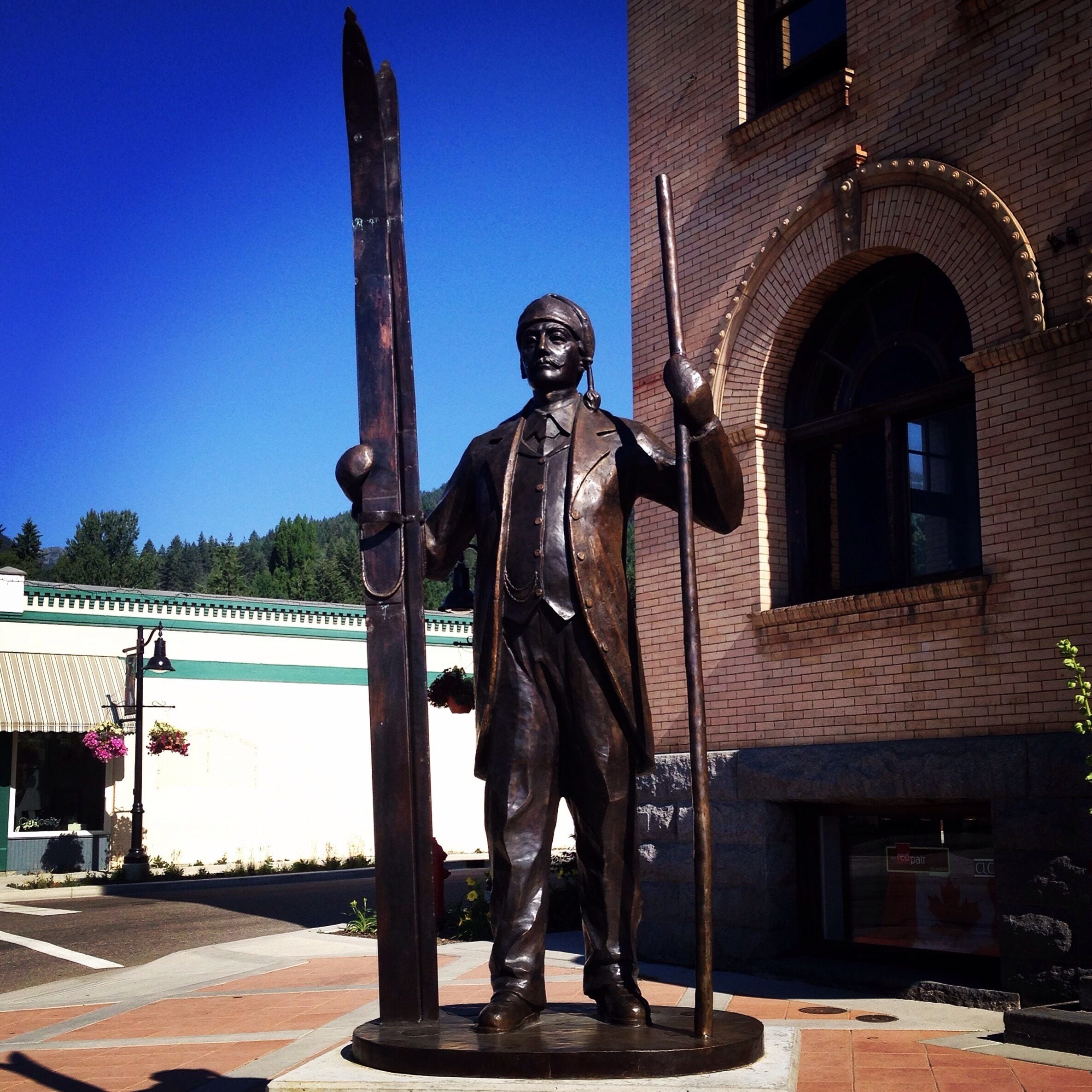 Uma estátua de bronze de um homem segurando uma espada e um cajado, em frente a um edifício de tijolos com toldo verde e torre do relógio.