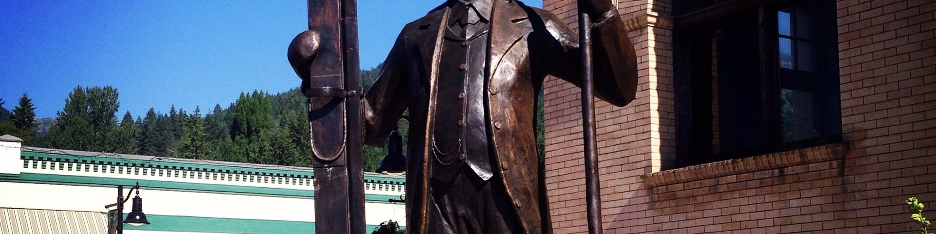 Uma estátua de bronze de um homem segurando uma espada e um cajado, em frente a um edifício de tijolos com toldo verde e torre do relógio.