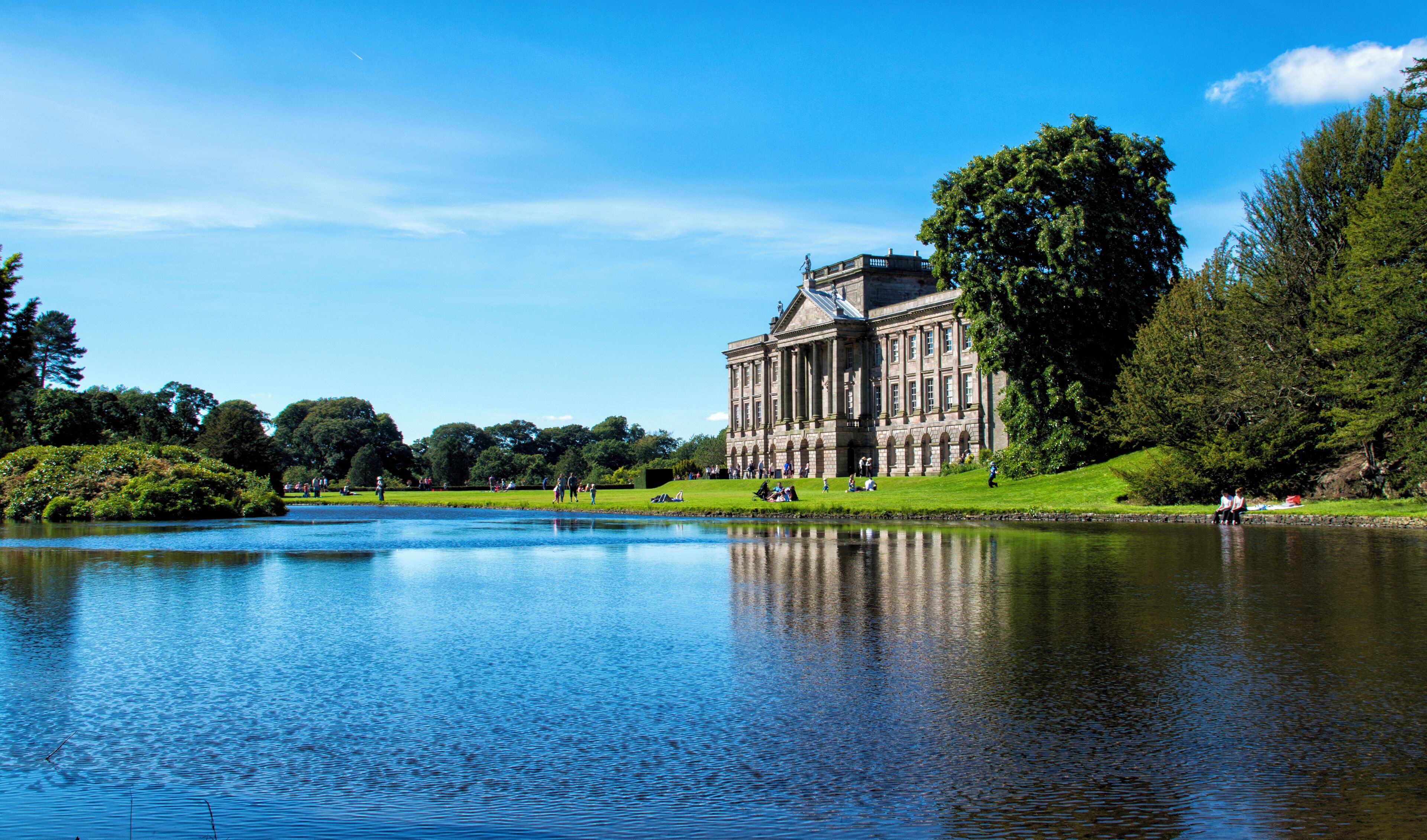 Um amplo edifício clássico com um lago espelhado, rodeado por vegetação exuberante e árvores.
