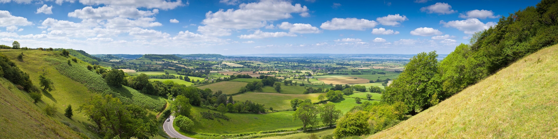 Vista panorâmica de um vale verdejante com colinas ondulantes, uma estrada sinuosa e uma cidade ao longe.